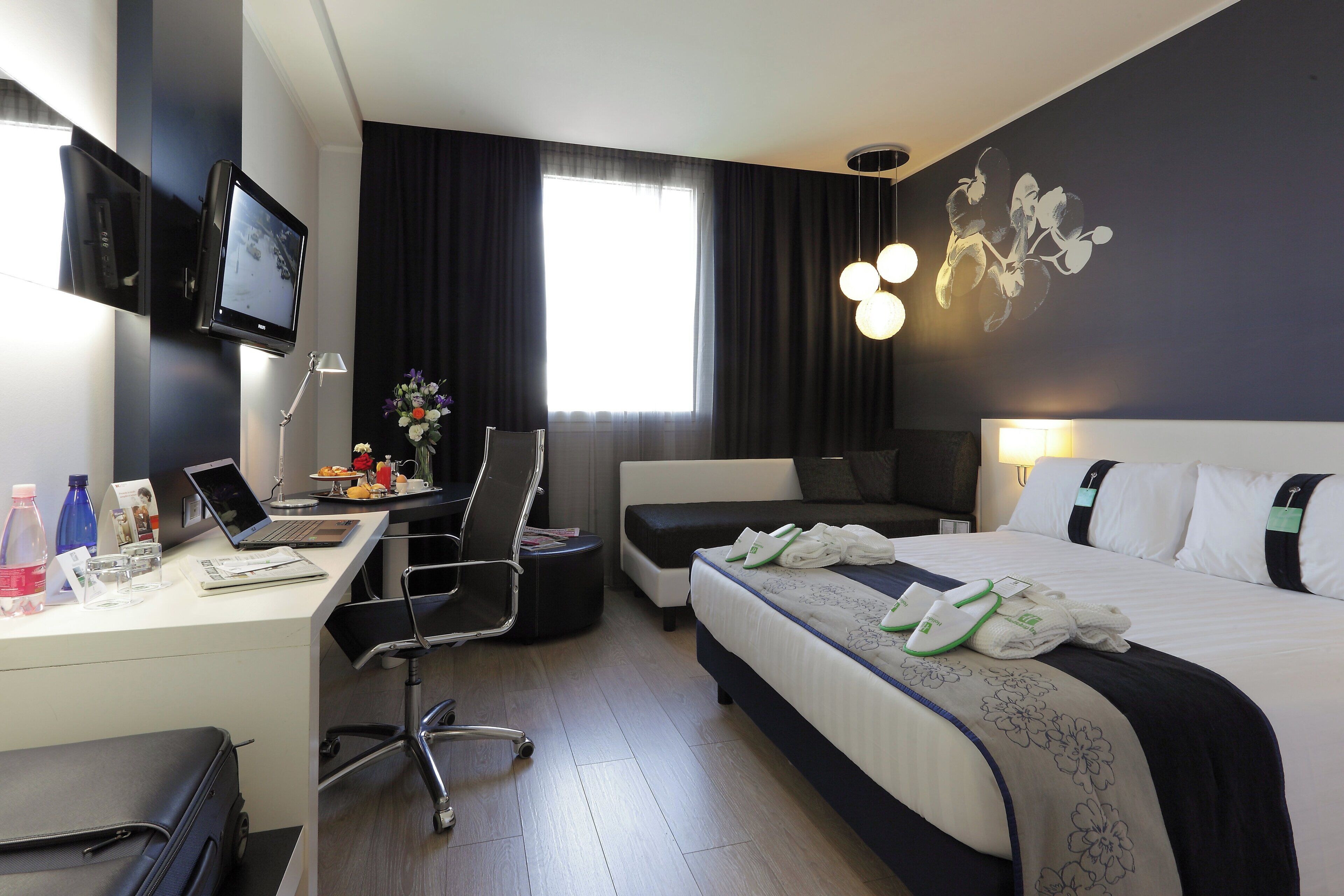 Quarto de hotel com escrivaninha, cadeira, cama e televisão fixada na parede.