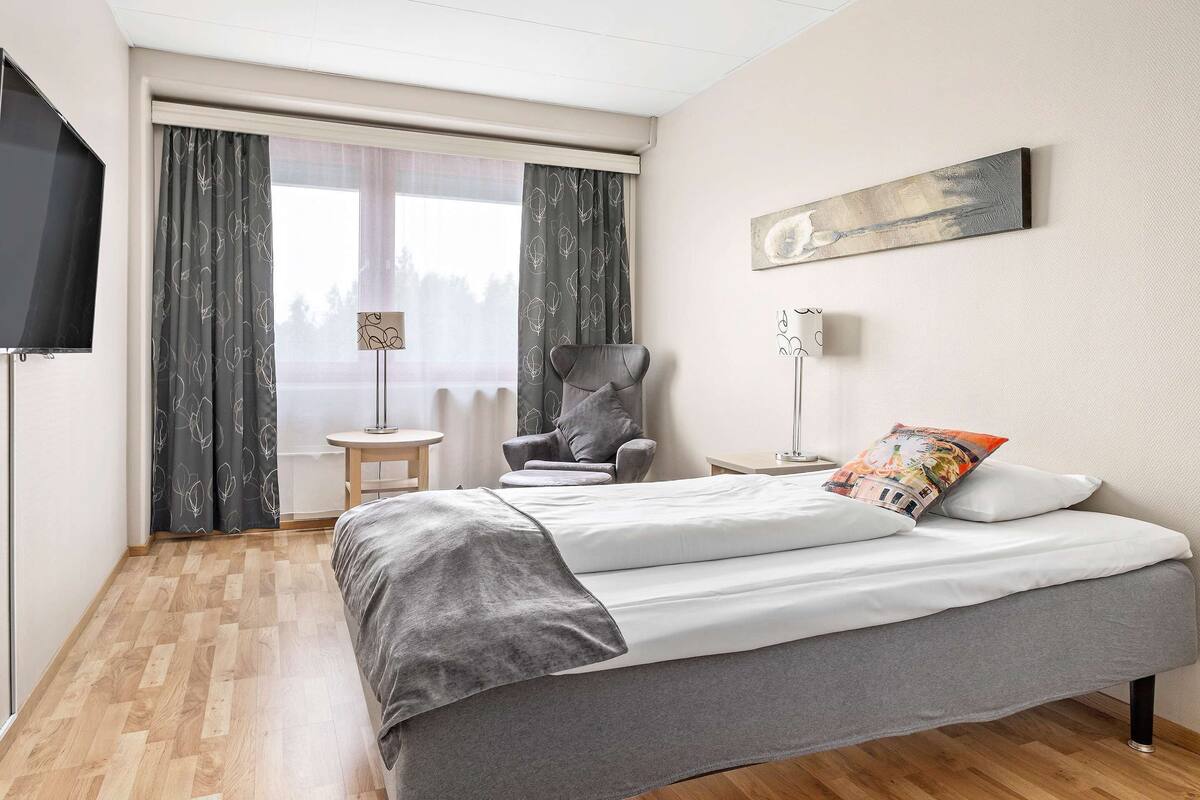 Quarto de hotel com cama, uma cadeira, mesa pequena, luminária e um quadro na parede.