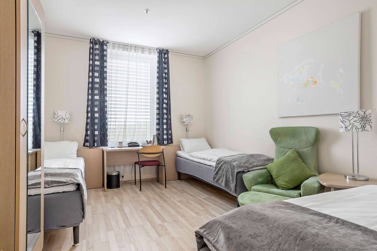 Quarto de hotel com uma cama, uma escrivaninha, uma cadeira e uma poltrona verde.