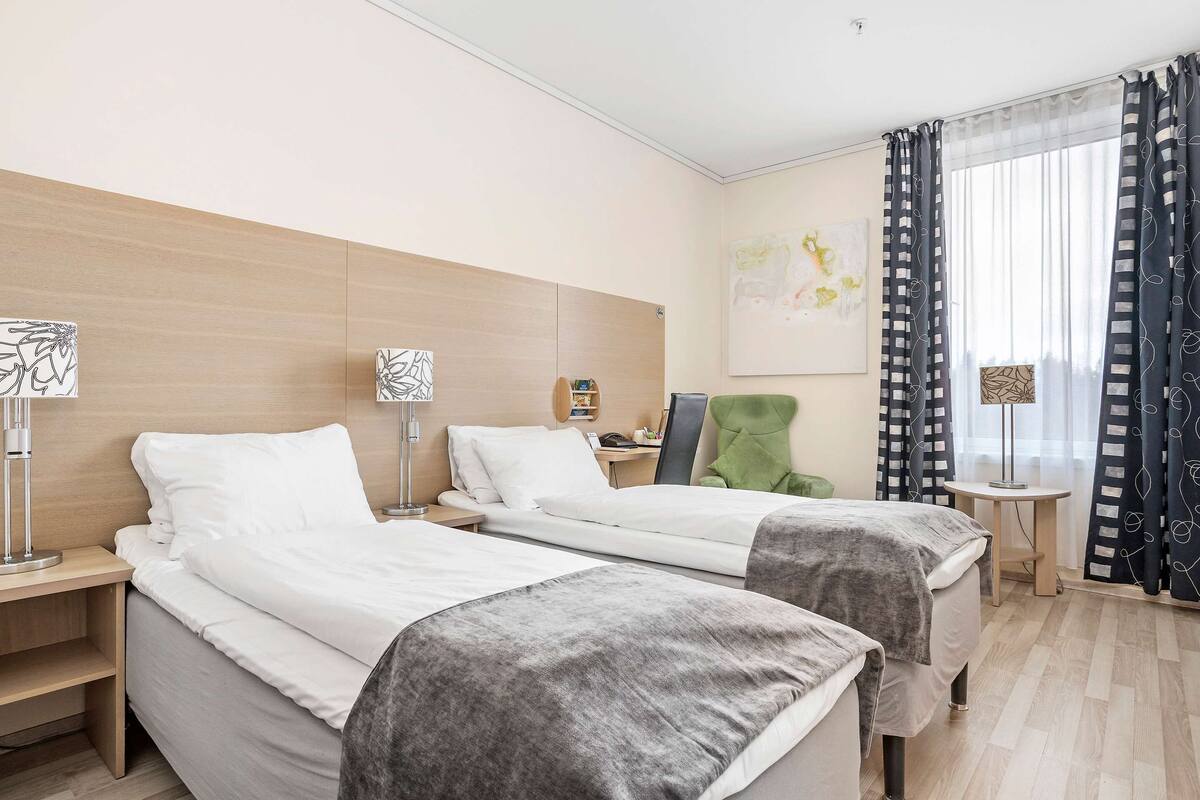 Quarto de hotel com duas camas, uma escrivaninha, uma cadeira, uma luminária e uma janela com cortinas.