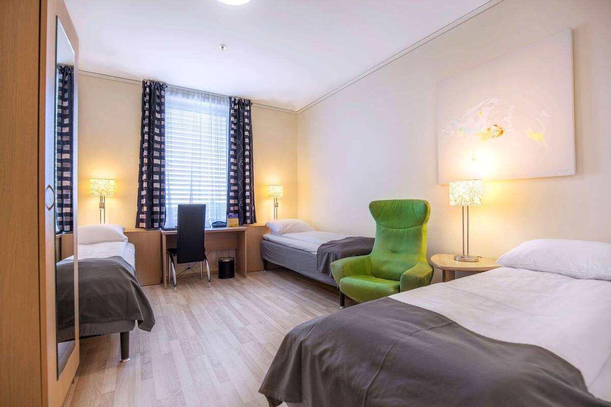 Quarto de hotel com duas camas, uma escrivaninha, uma cadeira verde e duas luminárias.