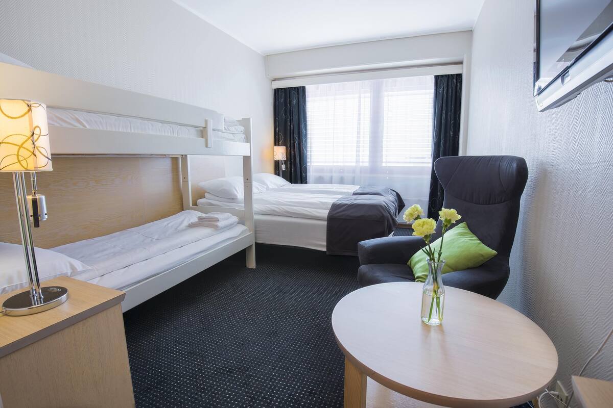 Quarto de hotel com cama beliche, cama de solteiro, escrivaninha com luminária, cadeira, mesinha e um vaso com flores.