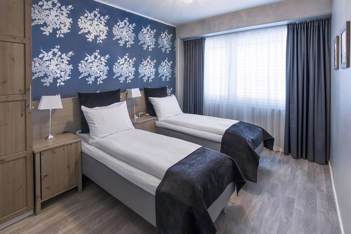 Quarto de hotel com duas camas, mesa de cabeceira com abajur, guarda-roupa de madeira, padrão floral na parede e janela com cortinas.