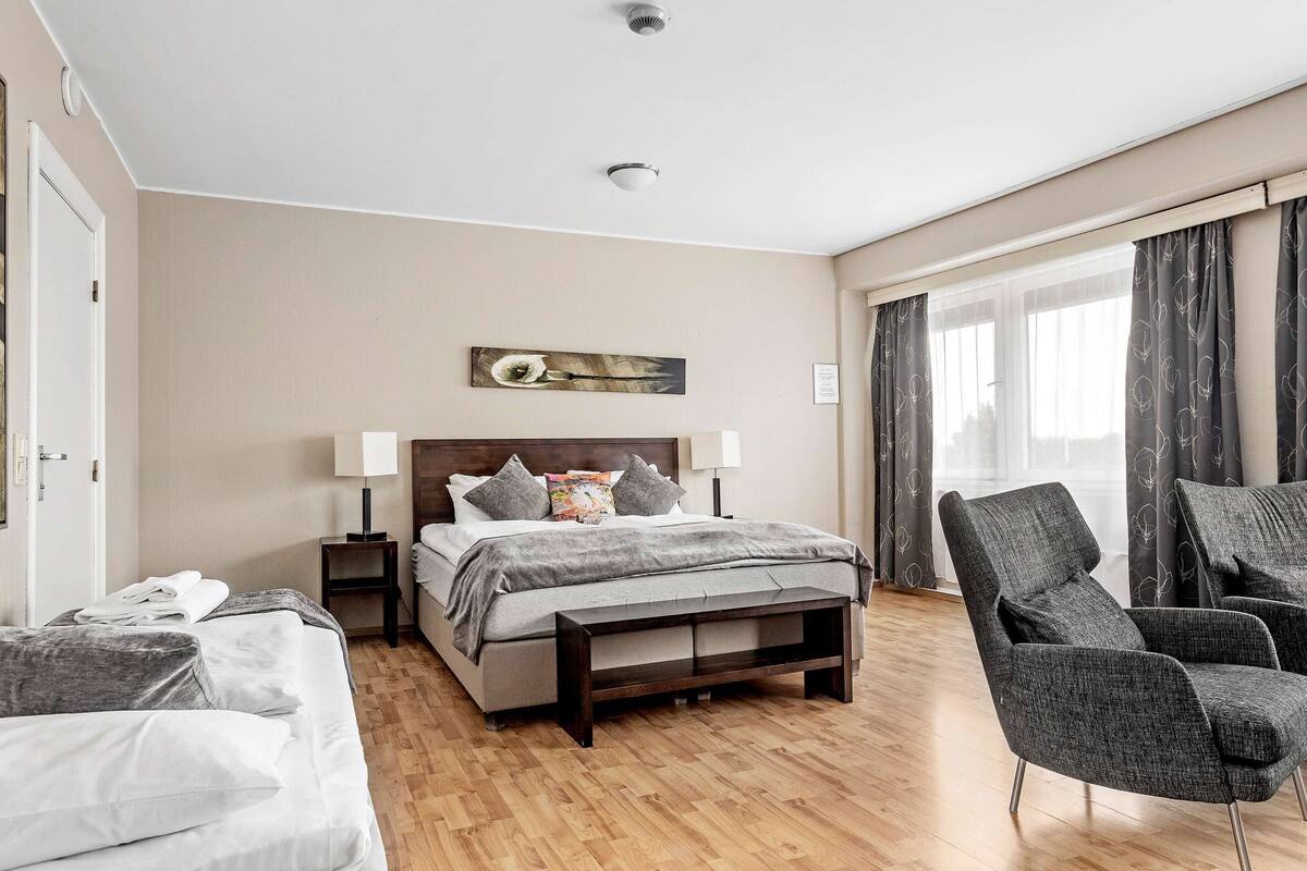 Quarto de hotel moderno com uma cama grande, mesas de cabeceira, um banco, uma cadeira e vista para o exterior pela janela.
