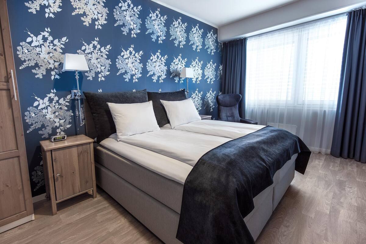 Cama bem arrumada com lençóis brancos, mesa de cabeceira com abajur e parede com padrão floral.