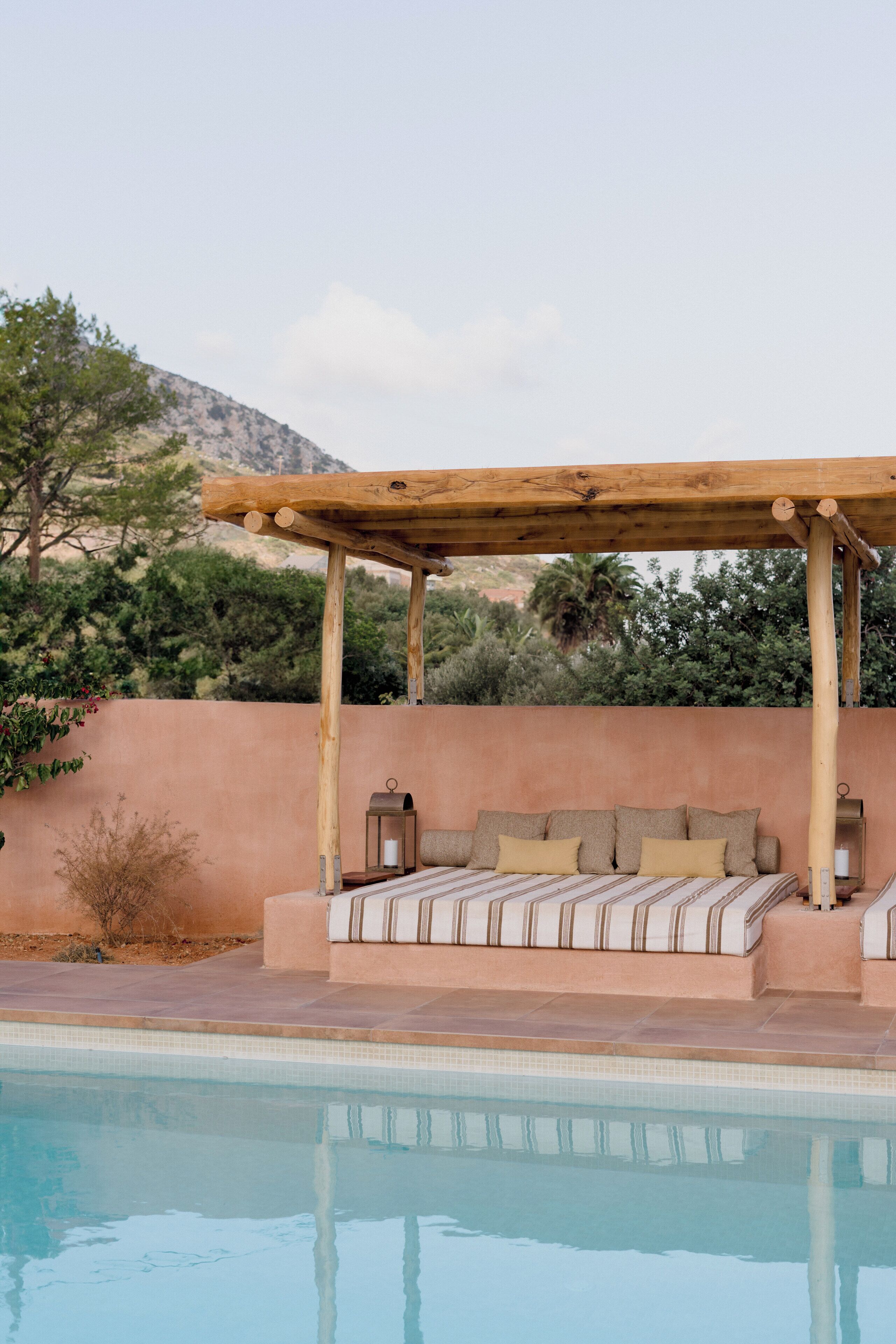 Uma área de estar à beira da piscina com um pérgola de madeira, bancos estofados e um candeeiro.
