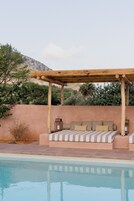 Uma área de estar à beira da piscina com um pérgola de madeira, bancos estofados e um candeeiro.