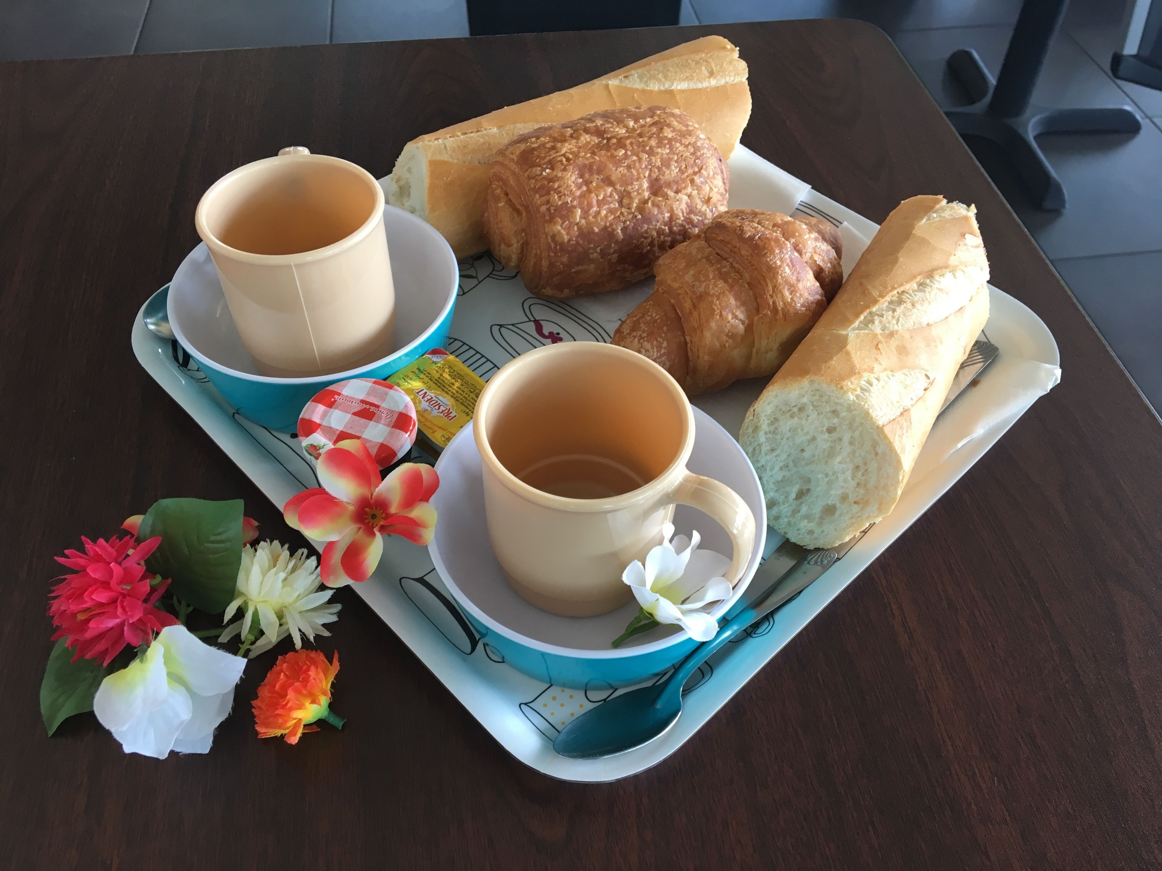 Uma bandeja com duas xícaras de café, dois croissants e um pequeno buquê de flores sobre uma mesa de madeira.