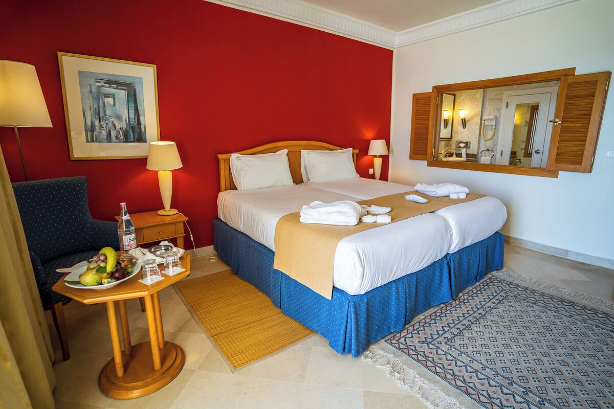 Quarto de hotel com uma cama, escrivaninha com uma fruteira, uma cadeira e um espelho.