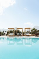 Uma área de piscina com espreguiçadeiras e pergolados de madeira, cercada por um edifício branco e palmeiras.
