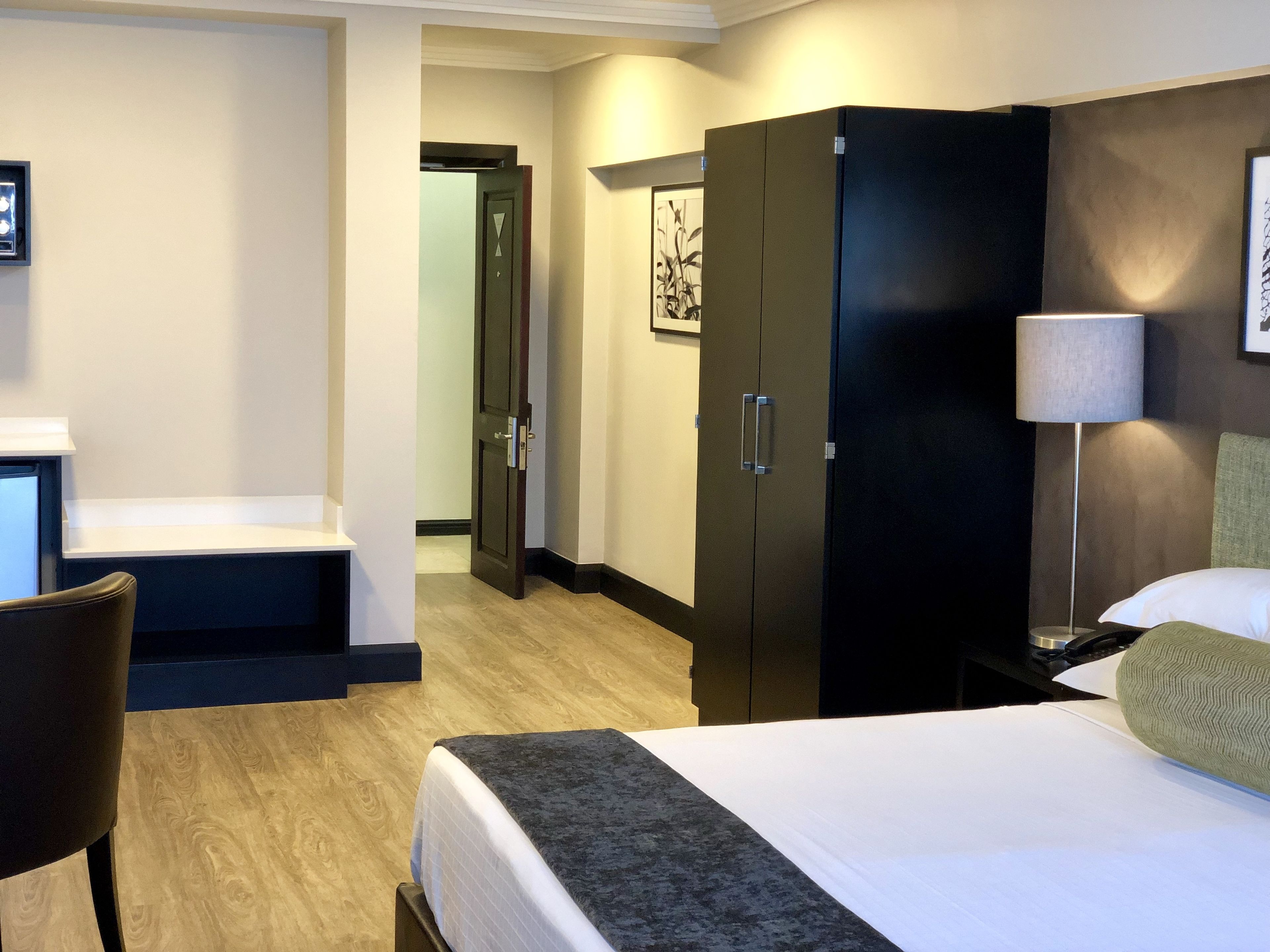 Quarto de hotel com cama, escrivaninha, cadeira, guarda-roupa e mesa de cabeceira com abajur.
