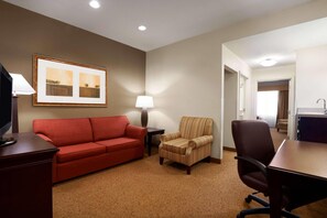 Quarto de hotel com sofá vermelho, poltrona listrada, mesinha de centro com abajur, televisão e escrivaninha de madeira.