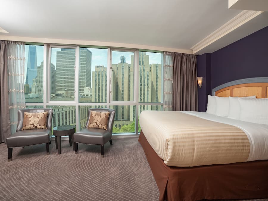 Quarto de hotel com uma cama grande, duas poltronas, uma mesinha e vista para edifícios da cidade através de grandes janelas.