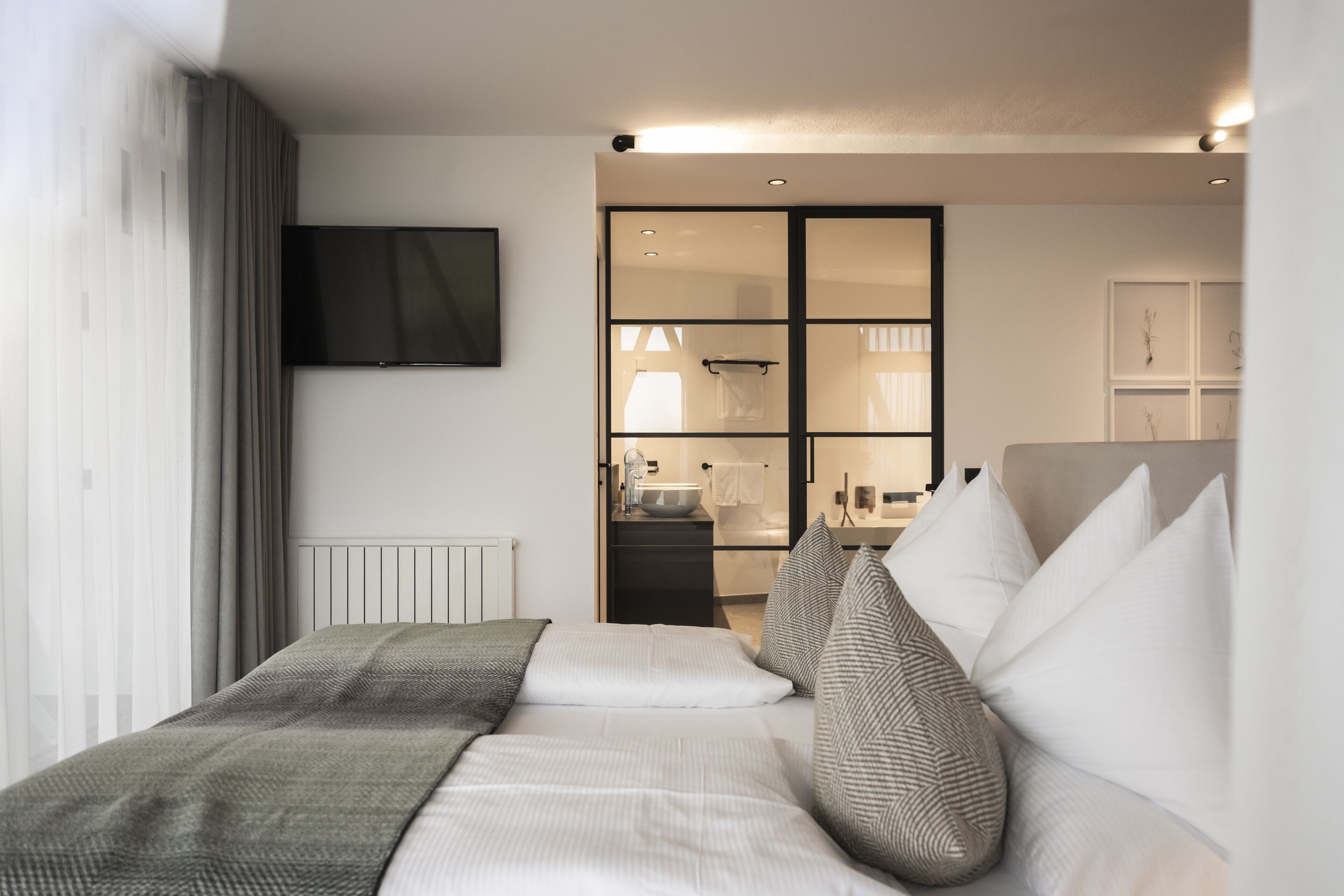 Quarto de hotel moderno com cama, televisão, banheiro visível por uma porta de vidro e janela com cortinas.