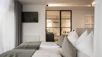 Quarto de hotel moderno com cama, televisão, banheiro visível por uma porta de vidro e janela com cortinas.