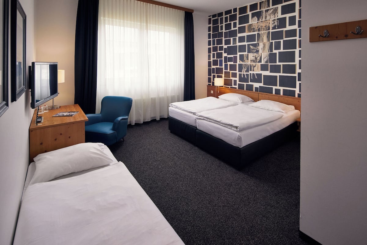 Quarto de hotel com duas camas, uma mesa de trabalho com televisão, uma cadeira e parede com padrão geométrico.