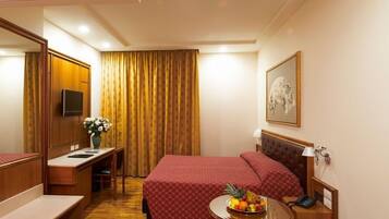 Quarto de hotel com cama, escrivaninha com computador, cadeira, pequena mesa com frutas e um quadro na parede.