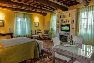 Quarto de hotel com cama, mesa de vidro, garrafa e copos, televisão, estante e uma cadeira.
