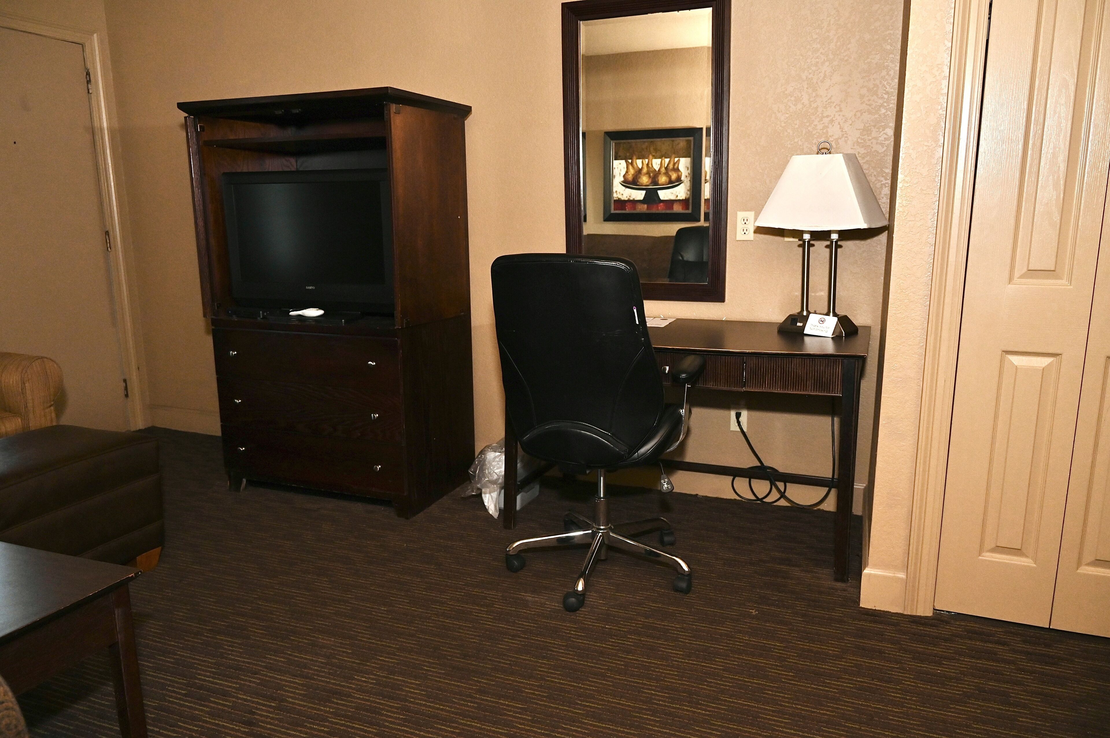 Quarto de hotel com televisão, secretária, cadeira, candeeiro e guarda-roupa.