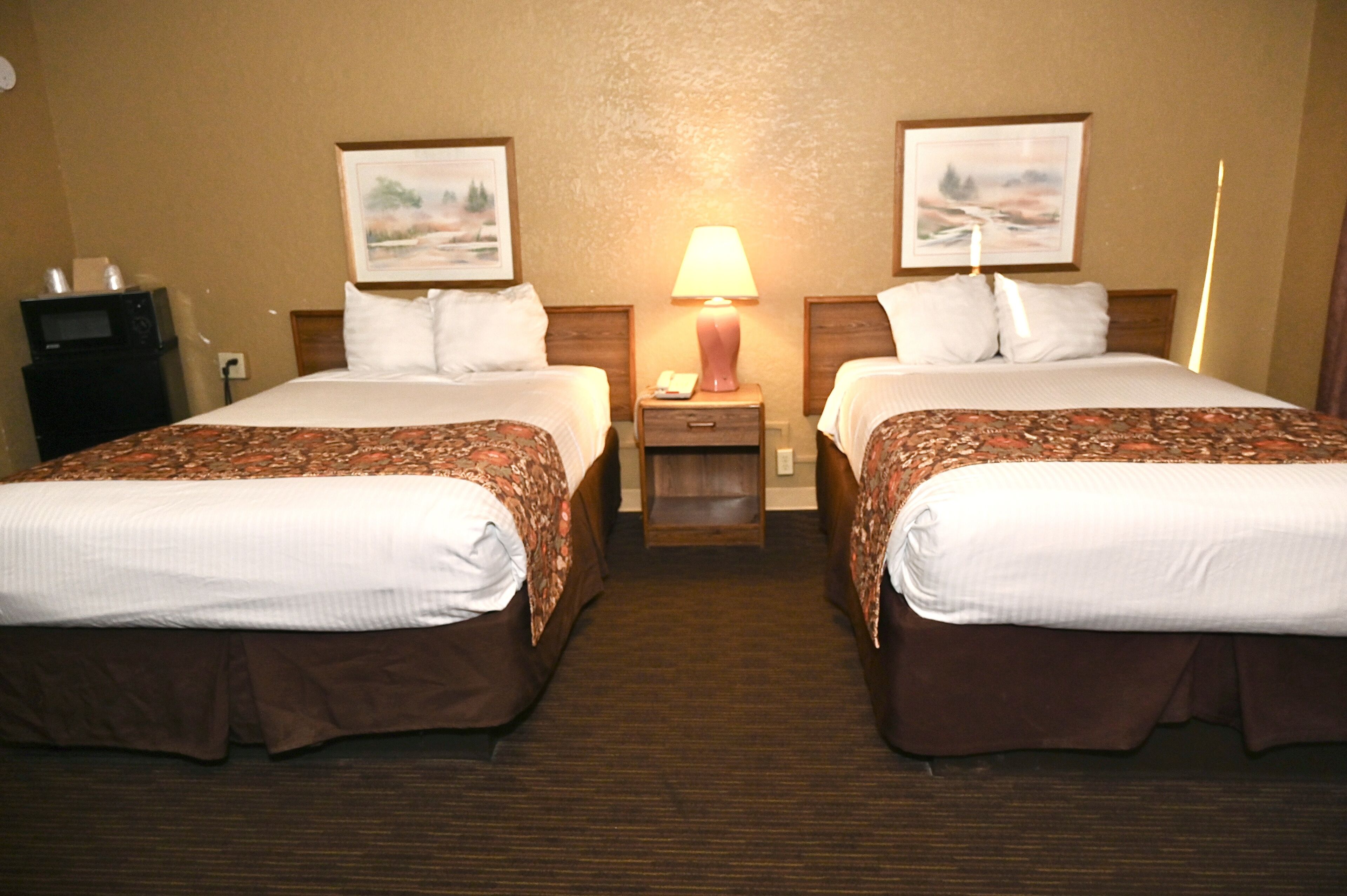 Quarto de hotel com duas camas, uma mesa de cabeceira com abajur e obras de arte nas paredes.