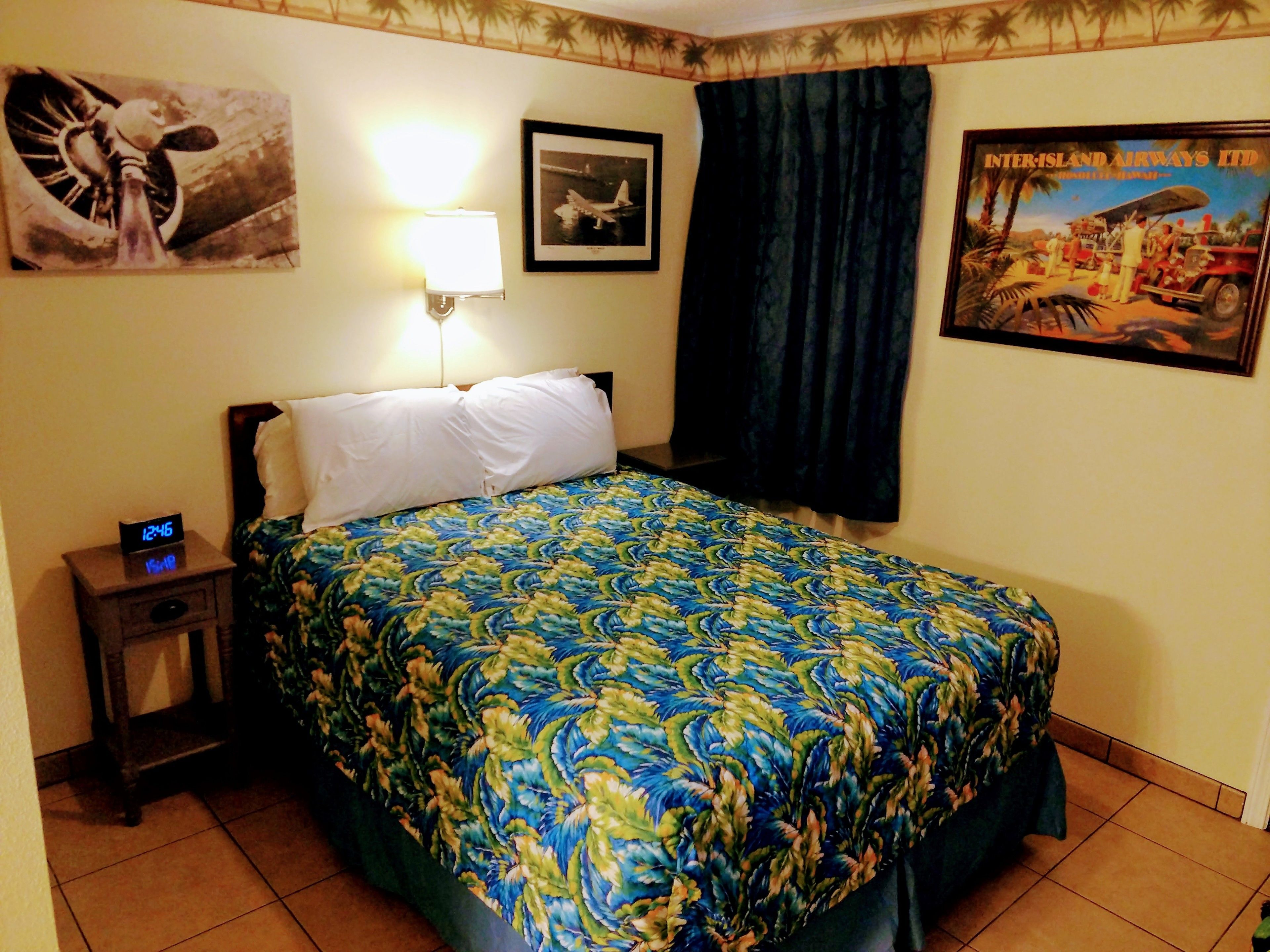 Quarto de hotel com cama, mesa de cabeceira com relógio, luminária na parede e quadros.
