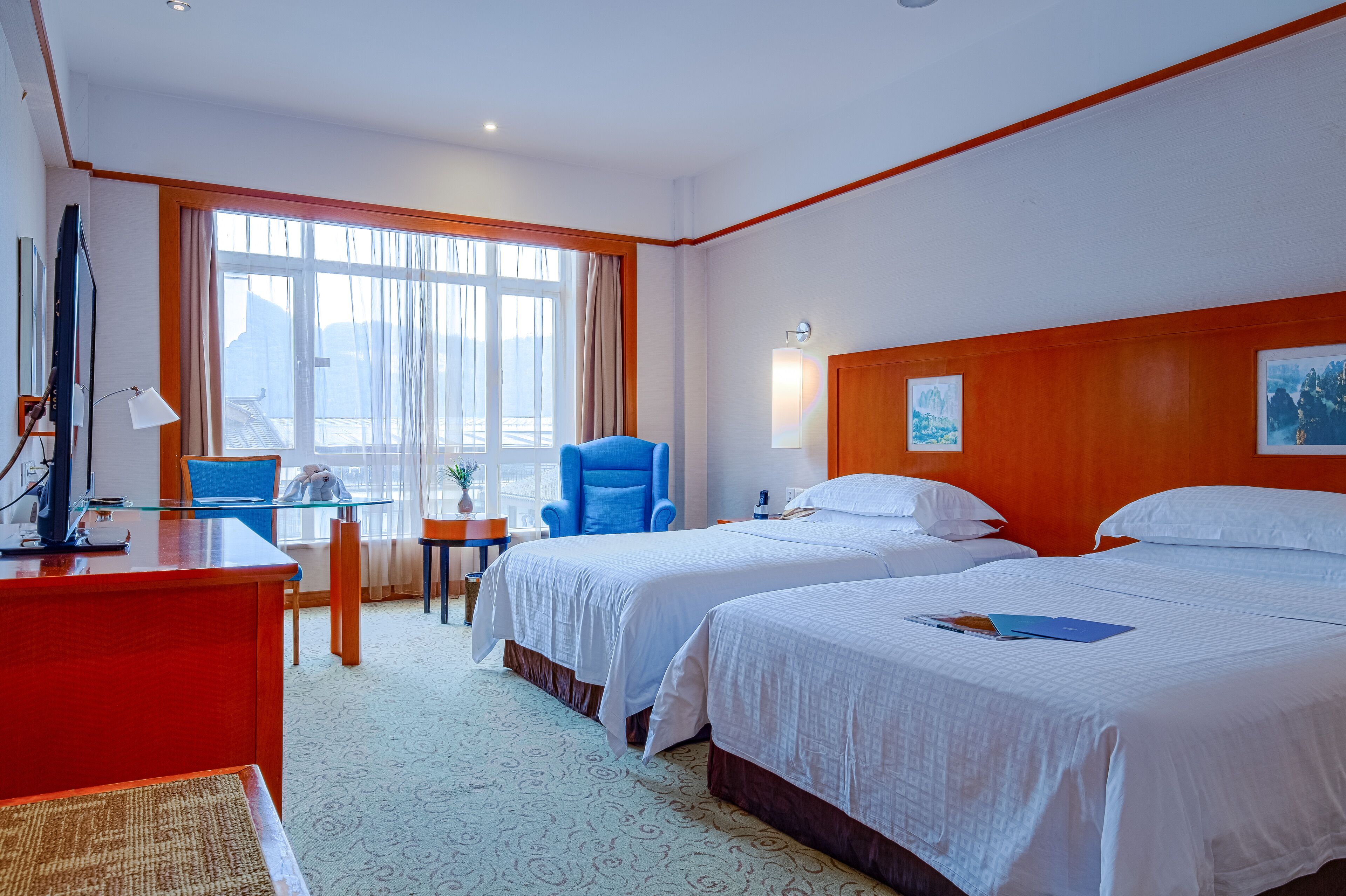 Quarto de hotel com duas camas, uma escrivaninha com computador, uma cadeira e uma janela com cortinas.