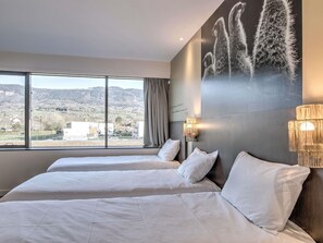 Quarto de hotel com duas camas, uma janela grande com vista para o campo, uma luminária na parede e um quadro.