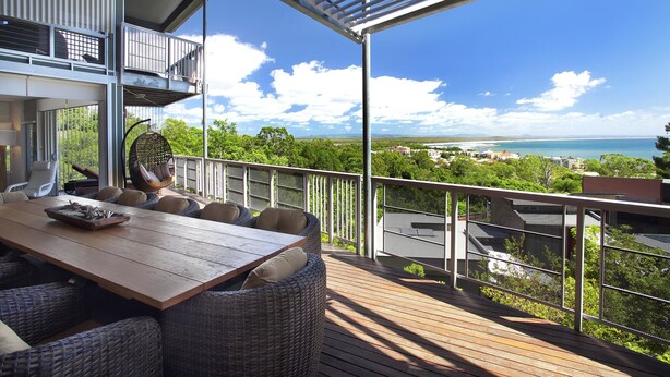 Um terraço com mesa de madeira e cadeiras de vime, com vista para uma paisagem deslumbrante, incluindo o mar e edifícios ao longe.