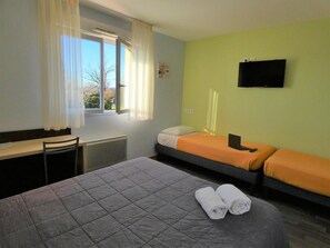 Quarto de hotel com cama, escrivaninha e cadeira. Janela com cortinas, televisão na parede e vista para o exterior.