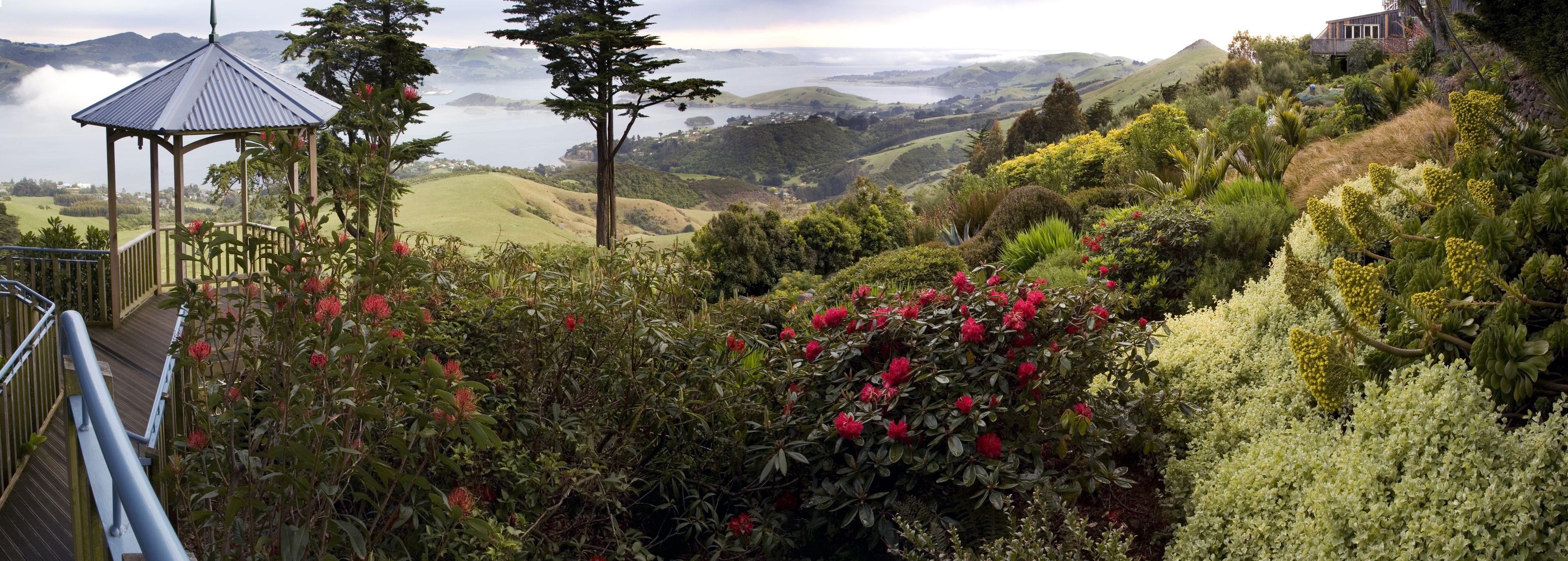 Um coreto com teto metálico, cercado por vegetação exuberante e plantas floridas.