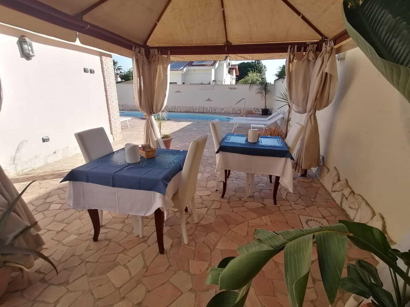Uma área de varanda coberta com duas mesas, cada uma com toalha e jarra. A varanda tem piso de azulejos e é cercada por paredes, com uma piscina ao fundo.