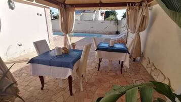 Uma área de varanda coberta com duas mesas, cada uma com toalha e jarra. A varanda tem piso de azulejos e é cercada por paredes, com uma piscina ao fundo.