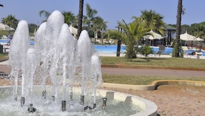 Uma fonte com vários jatos de água em área ajardinada, com palmeiras e uma piscina ao fundo.
