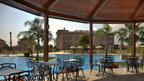 Área à beira da piscina com mobiliário de exterior e vista para um edifício grande ao fundo.