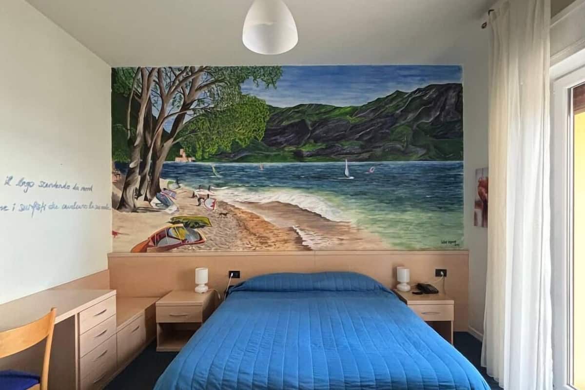 Quarto com um mural grande mostrando uma cena de praia, uma cama com colcha azul, uma escrivaninha e uma cadeira.