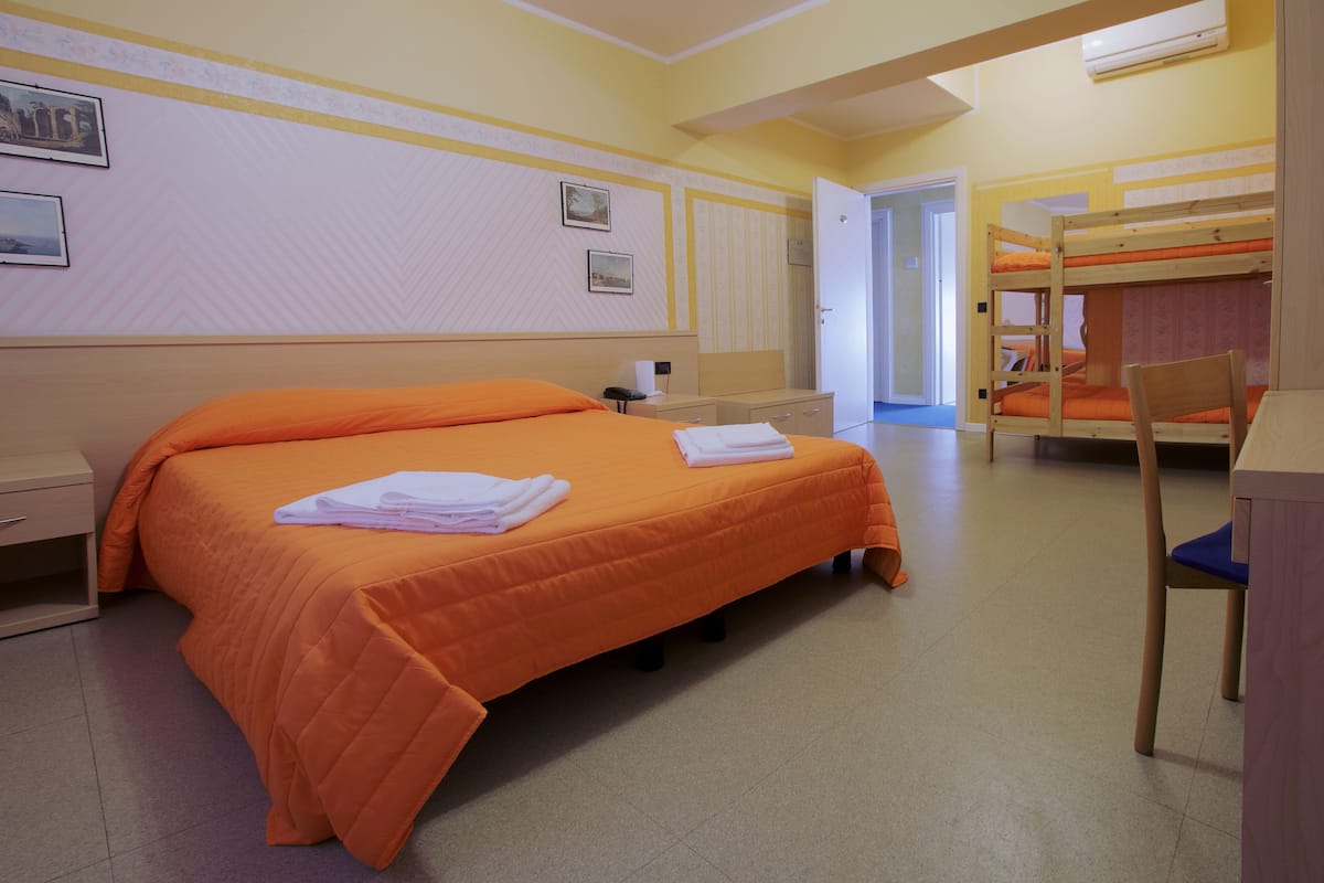 Quarto de hotel com uma cama de solteiro, uma mesa, uma cadeira e uma cama beliche ao fundo.