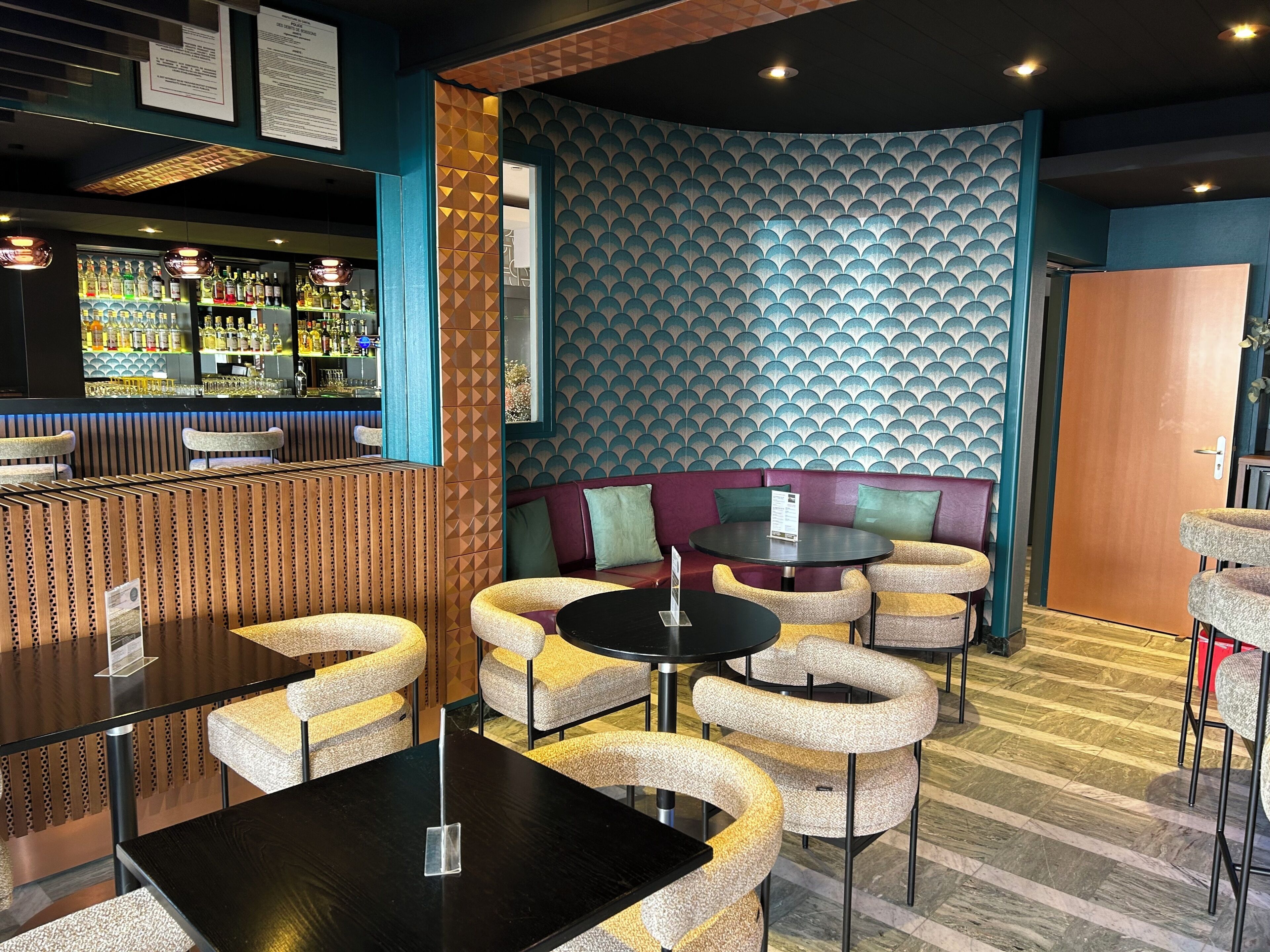Uma área de bar com balcão exibindo diversos copos e garrafas, espaço de estar com mesas redondas e cadeiras, e uma parede com padrão.