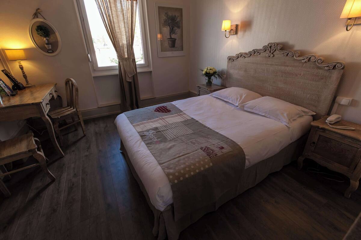 Um quarto bem arrumado, com cama, mesa de cabeceira, abajur, cadeira e escrivaninha.