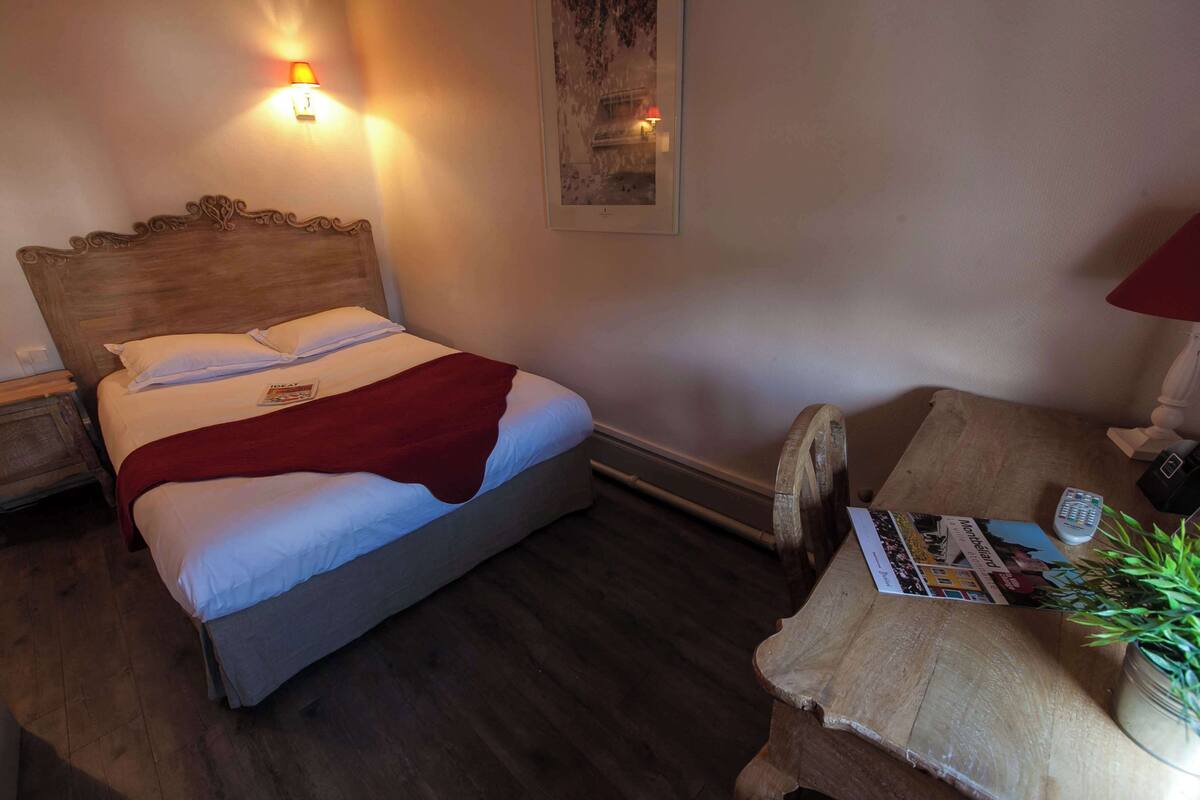 Uma cama bem arrumada com manta vermelha, cabeceira de madeira e luminária fixada na parede.