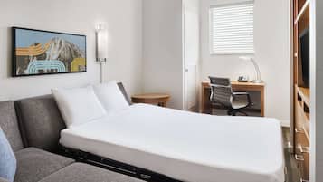 Quarto de hotel com cama, escrivaninha, cadeira, mesa redonda pequena e um quadro na parede.
