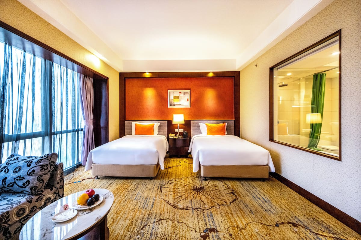 Quarto de hotel com duas camas, um sofá, uma mesa de jantar com frutas e uma janela grande com cortinas.