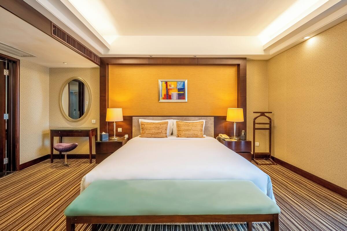 Quarto de hotel com uma cama grande, duas mesas de cabeceira com luminárias, um espelho redondo, um banquinho pequeno e uma obra de arte na parede.