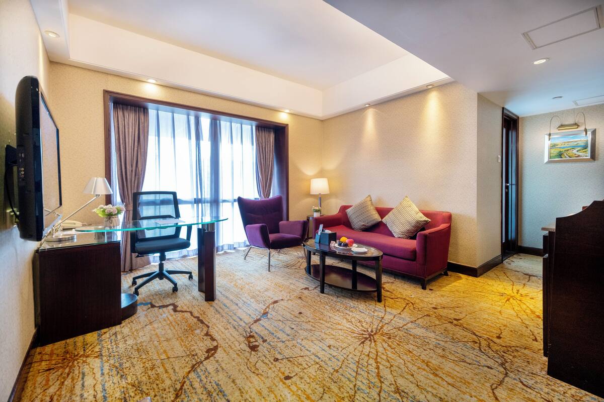 Quarto de hotel com escrivaninha de vidro, sofá vermelho, poltrona roxa e mesa de centro.