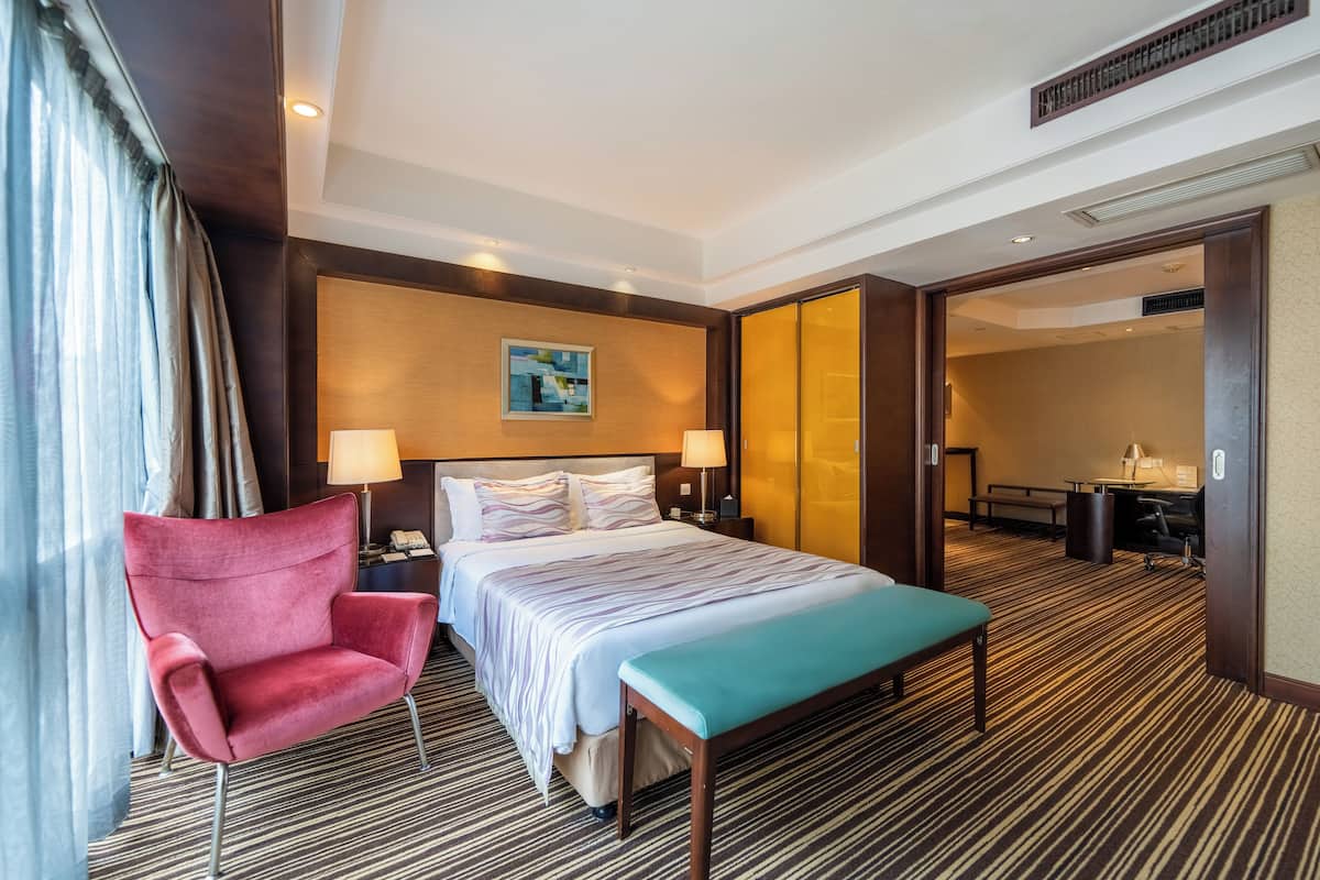 Quarto de hotel com cama, poltrona, banco, escrivaninha e banheiro visível por uma porta aberta.