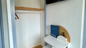 Quarto compacto com cama, uma pequena mesa com banquinhos, TV fixada na parede e prateleira de madeira.
