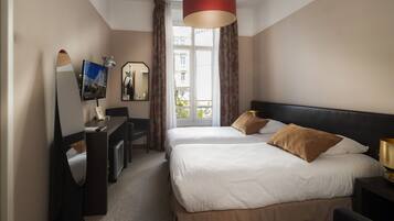 Quarto de hotel com duas camas, uma escrivaninha, uma cadeira, televisão e uma janela grande com cortinas.