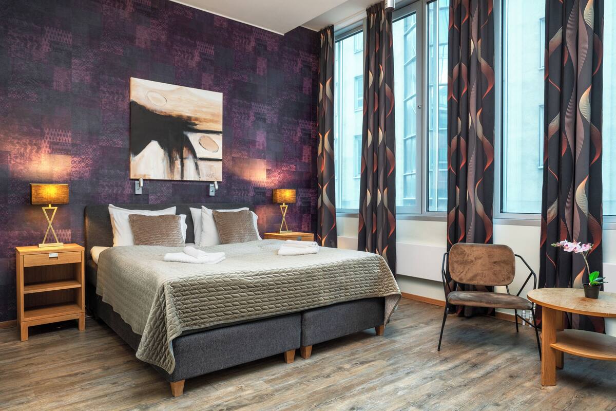 Quarto de hotel moderno com uma cama grande, duas mesas de cabeceira com luminárias, uma cadeira e uma pequena mesa com uma planta.