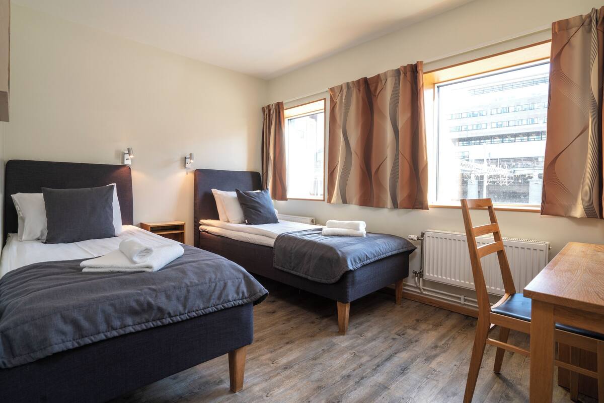 Quarto de hotel com duas camas, uma mesa de madeira e vista para um edifício através de grandes janelas.