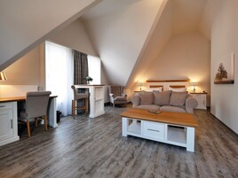 Quarto moderno estilo loft com teto inclinado, mesa de centro de madeira, sofá, escrivaninha com cadeira e pequena mesa com abajur.