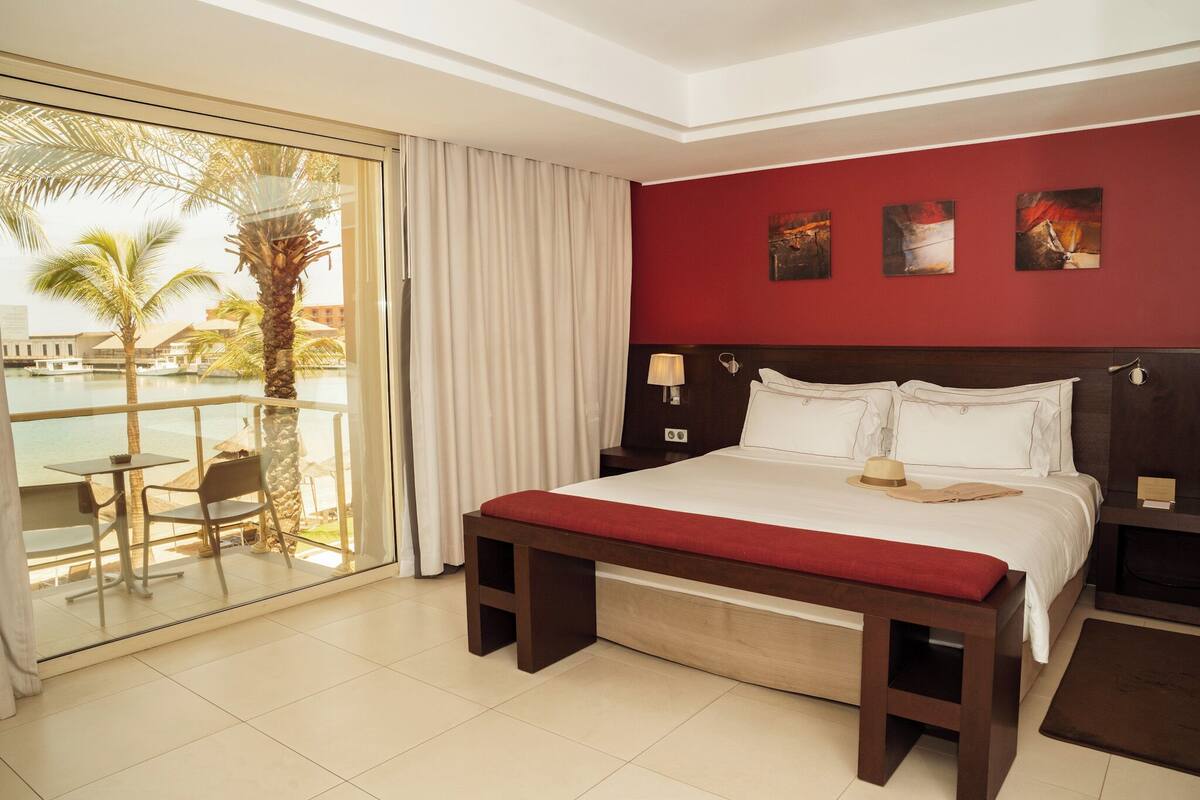 Quarto de hotel com uma cama grande, uma parede de destaque vermelha e vista para uma piscina e palmeiras através de uma porta de vidro.